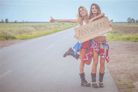 Two Beautiful Cheerful Hipster Friends Hitchhiking By The Road Go
