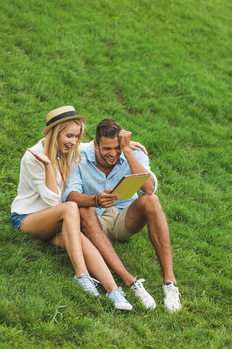 Cheerful Couple Using Tablet Together While Sitting On Green Lawn