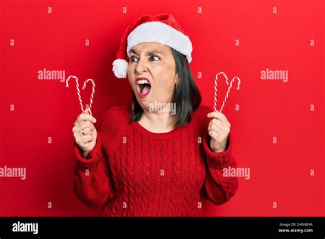 Middle Age Hispanic Woman Wearing Christmas Hat Holding Candy Angry And