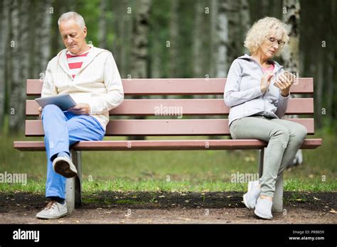 Senior Man And Woman Sitting On Opposite Sides Of Bench And Using