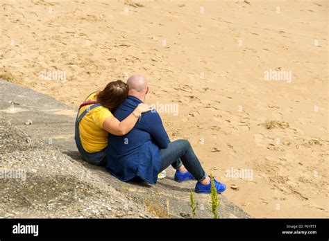 A Couple Sitting On A Rock Above A Sandy Beach The Woman Has Her Arm