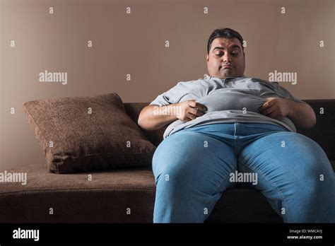 Obese Man Sitting On Sofa Holding His Belly Fat With Both Hands And