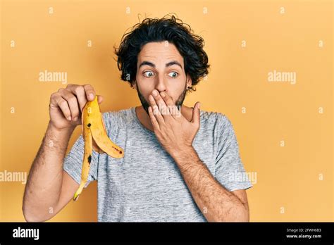 Handsome Hispanic Man Holding Banana Peel Covering Mouth With Hand