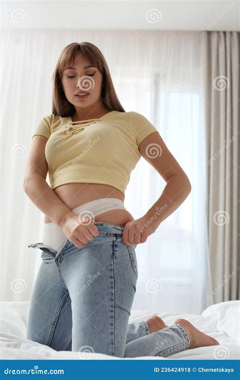 Young Woman Struggling To Squeeze Into Tight Jeans On Bed At Home Stock