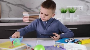 Education, school, learning difficulties, reading difficulties. Child plays with smartphone instead of doing homework. Schoolchild sits at table doing homework among pile of books and scattered toys.