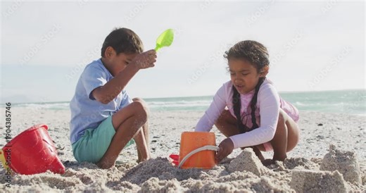Happy hispanic brother and sister playing on beach with sand and building sand castle