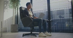 Side view of hardworking Chinese manager or freelancer working at his portative laptop. Man sitting at chair near large panoramic windows with drapes. Distracted from work while looking outside.