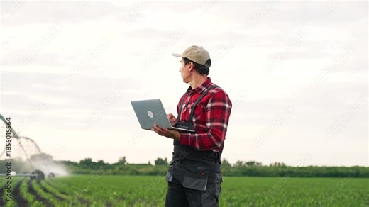 Farmer checks laptop in field using agriculture technology for irrigation and crop inspection near pivot and tractor on farm wearing cap and overalls while monitoring irrigation and crop health