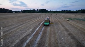 Tractor collects square bales of straw with bale picker from a recently harvested field and places them on the transport wagon in the evening.