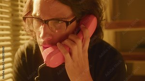 Young man in eyeglasses speaking on retro pink telephone, sitting on window sill in living room with warm light
