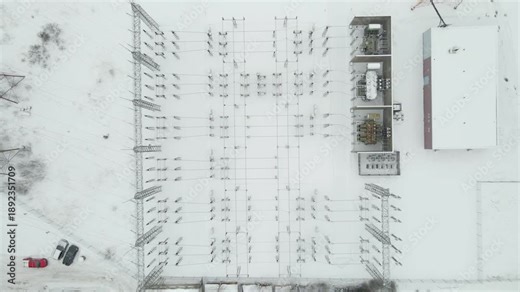 High altitude top down aerial view of a snow covered electrical substation, revealing full power grid layout as the camera slowly descends for a cinematic overview.