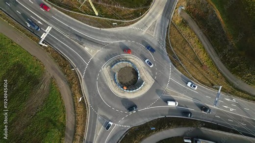 Drone shot looking straight down at a multi-lane roundabout with cars and trucks circulating smoothly, surrounded by rural roads and fields.