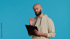 Middle eastern adult creating a checklist on clipboard papers, being focused on trying to plan tasks on an organizer in studio. Modern person looking at files and listing things to do. Camera 1.