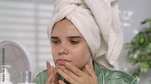 A young woman squeezes a pimple on her chin in front of a mirror. A woman after a shower, wearing a white bath towel over her head and green silk pajamas, examines her face in a close up mirror.