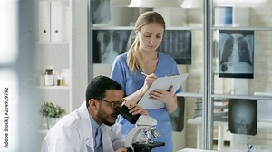 Tilt down shot of young black man looking into microscope in laboratory and discussing work with Caucasian colleague who is writing data into document attached to clipboard