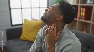 A relaxed african american man scratches his neck while sitting on a couch indoors, with a bookshelf in the background