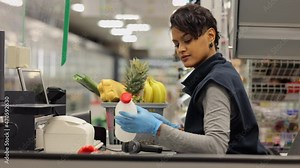 Young woman cashier scanning fresh food and doing work at checkout of organic store spbi. Closeup view of African American female takes milk, eggs from basket and scans in front of computer screen