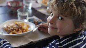 Young boy turning head toward camera seated at lunch table smiling. Child eating pasta food, turns head back to plate. Handsome portrait