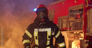 African American young brave fireman in the equipped costume, helmet and gas mask walking and rushing from a fire truck to fight with a fire. At night.