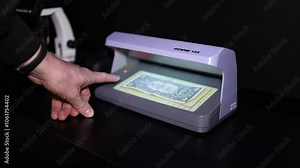 A person uses a banknote verification machine, placing a bill under the UV light to check its authenticity. The bright light highlights security features, ensuring the note is genuine. This setup is i