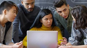 Dollying waist-up shot of young mixed race female creative professional sitting at table and typing document on laptop, with input and suggestions from her colleagues, who are standing and watching