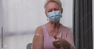 Senior woman wearing face mask showing vaccinated arm with adhesive bandage plaster and gesturing thumbs up. Happy elderly patient approving vaccination. Covid-19 Vaccination program concept