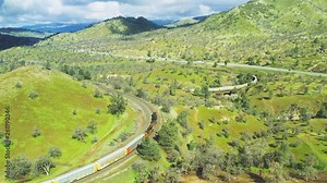 The aerial camera follows a 5000-foot-long freight train pulled by three massive diesel locomotives around the Tehachapi Railroad Loop with California Highway 59 in the background.