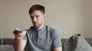 Caucasian man tries to press buttons on TV remote control while sitting on sofa. Non working device and negative emotions from unsuccessful attempts to turn on device with infrared remote control.