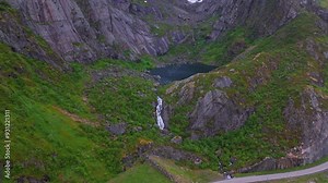 Olkonvatnet near Reine in Lofoten Norway. Small snow melting overflow pond on a mountain range, with a waterfall streaming down towards the sea. Mountain peaks on all sides, green vegetation on ground