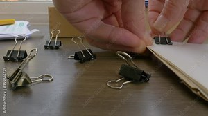 Closeup POV dolly shot past sprung metal clips (bulldog clips / springback clips / binder clips), to a man’s hands using one to clamp some documents together.