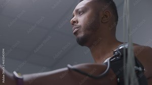 African American man with tonometer and sensors pedals orbitrek in cardiology room during holter monitoring procedure. Patient with cardiovascular disease. Electrocardiography in hospital. Portrait.