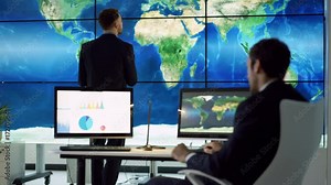 Businessmen discussing large world map graphic on video wall, with pie charts & graphs displayed on computer screen. Global business concept.