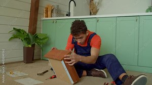 Concentrated handsome black handyman in protective workwear completing assembly of DIY bedside table drawer, adjusting front wooden panel while sitting on floor in domestic room.