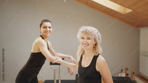 Pilates lesson on chair, side view of blond elderly personal trainer coaching two young women indoor at studio. Correction of musculoskeletal system, beautiful body. Correct posture. Sun glare effect