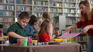 Joyful female teacher and little girl with autism stretching slime to inflate bubble during masterclass for kids with disabilities. Healthy children and kids with special needs spending time together