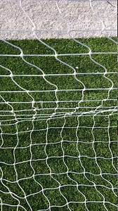 Looking down at a white soccer goal netting as part of a soccer backstop net structure. The sports netting waves in the wind Set against green and white turf athletic field.