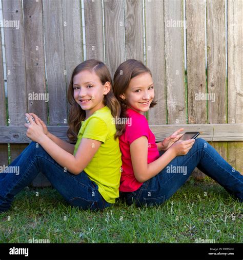 Twin Sister Girls Playing With Tablet Pc Sitting On Backyard Lawn Fence Leaning On Her Back