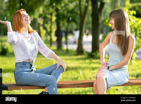 Two Young Girls Friends Sitting On A Bench In Summer Park Chatting Happily Having Fun Stock