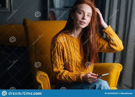 Portrait Of Gorgeous Redhead Young Woman Using Smartphone Sitting In Yellow Armchair Smiling