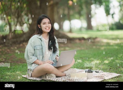 Asian Woman Sitting On Picnic Blanket And Lawn At Park Working On Laptop Asian Female Using