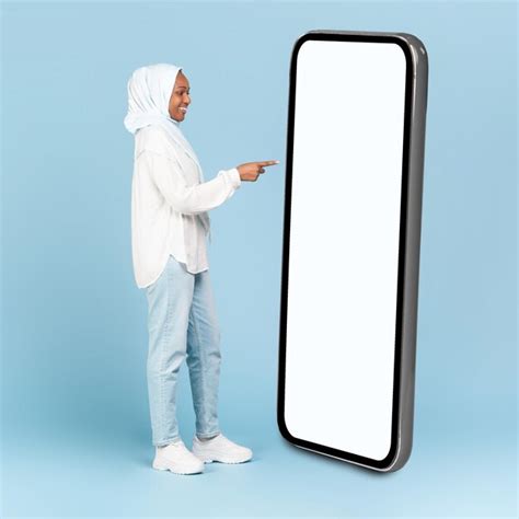 空の白い画面で大きなスマートフォンを使用しディスプレイのモックアップを指している黒人のイスラム教徒の女性の全身の長さ プレミアム写真