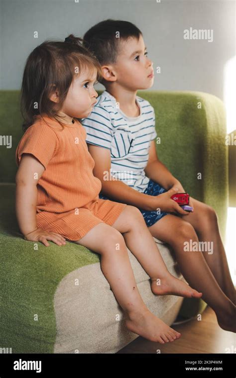 Excited Small Brother And Sister Sitting On Sofa Watching Tv In Living Room Relaxing At Home On