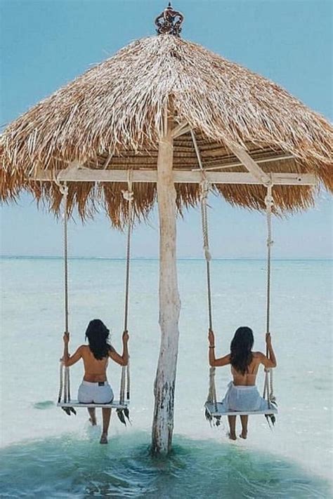 Two Women Sitting On Swings In The Ocean Under A Thatch Roof Over Looking The Water