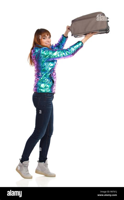 Young Woman In Vibrant And Shiny Down Jacket Puts The Luggage Up Looks At Camera And Smiles