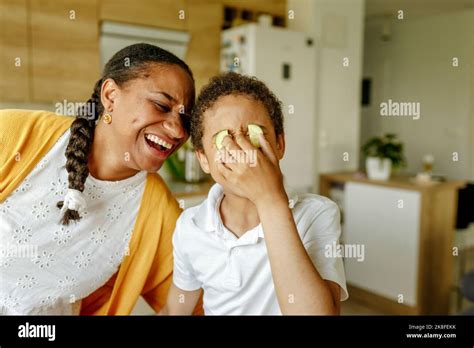 Happy Mother Embracing Son Covering Eyes With Cucumbers At Home Stock