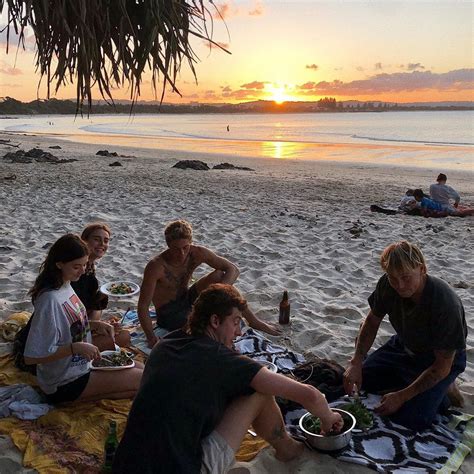 A Group Of People Sitting On Top Of A Beach Next To The Ocean At Sunset