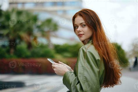 Woman With Phone Walking Down The Street In The City In Fashionable Clothes With Red Hair And A